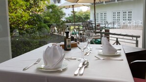Mesa posta para refeição com toalhas brancas, talheres e taças de vinho, com vista para um jardim e edifícios.