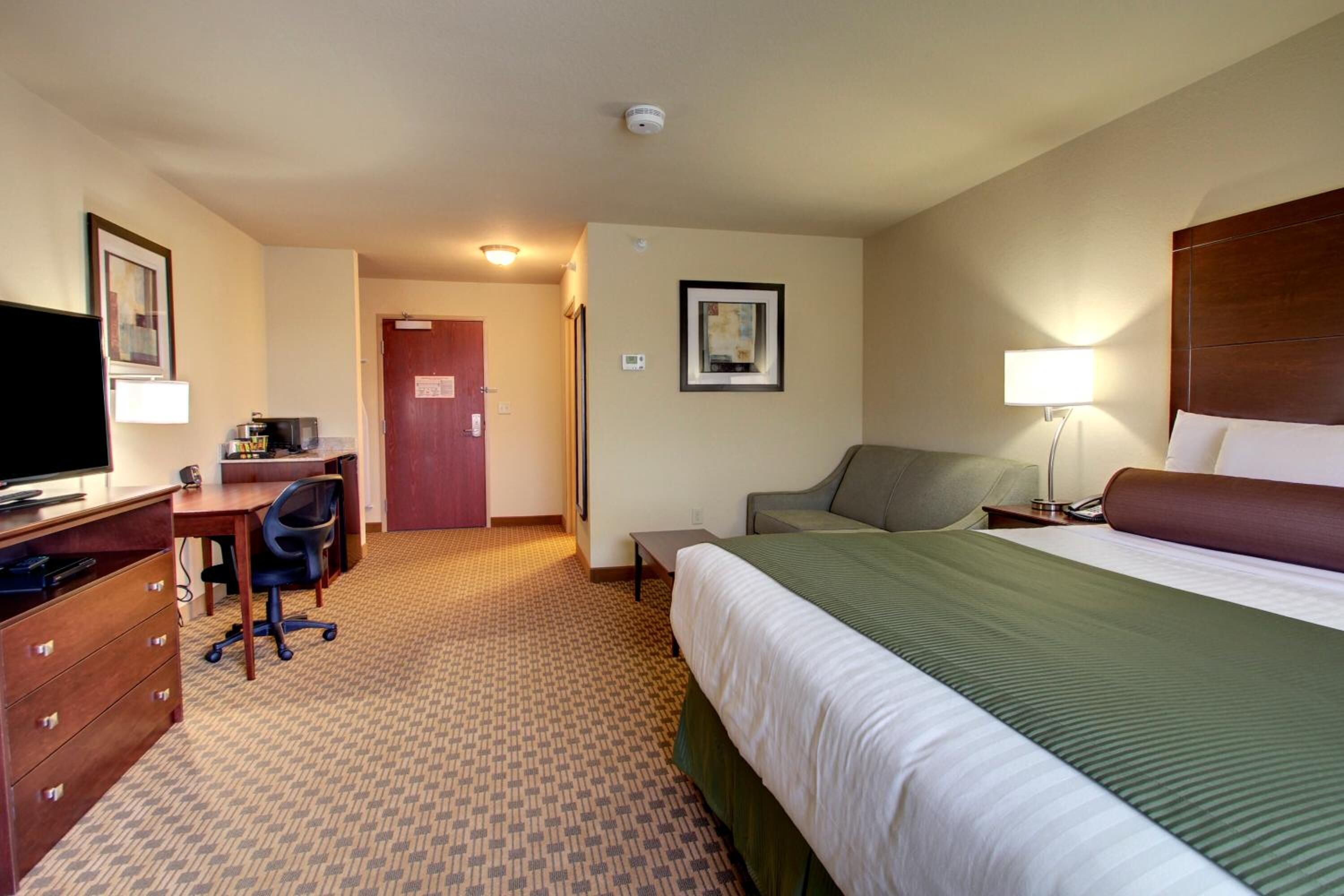 Quarto de hotel com cama, escrivaninha, cadeira, televisão e um quadro na parede.