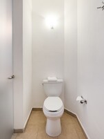 Casa de banho pequena e branca, com sanita, rolo de papel higiénico preso à parede e porta.