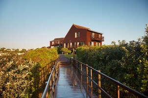 Uma passarela de madeira leva a uma casa grande e moderna com varanda, rodeada por vegetação exuberante.