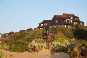 Uma casa de madeira grande, com vários andares, e uma escadaria que leva a um deck, cercada por vegetação densa e dunas de areia.