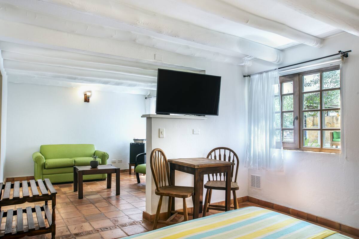 Quarto com sofá verde, mesa de jantar de madeira, cadeiras, televisão na parede e janela com cortinas.