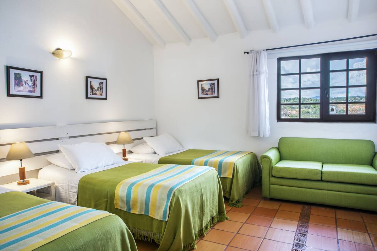 Quarto de hotel com duas camas, um sofá verde, uma janela com vista e quadros nas paredes.