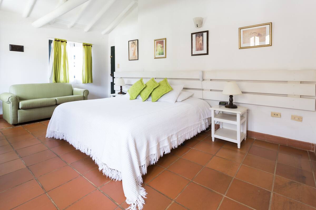 Quarto com cama, almofadas verdes, mesa de cabeceira branca, luminária, sofá verde e janela com cortinas.
