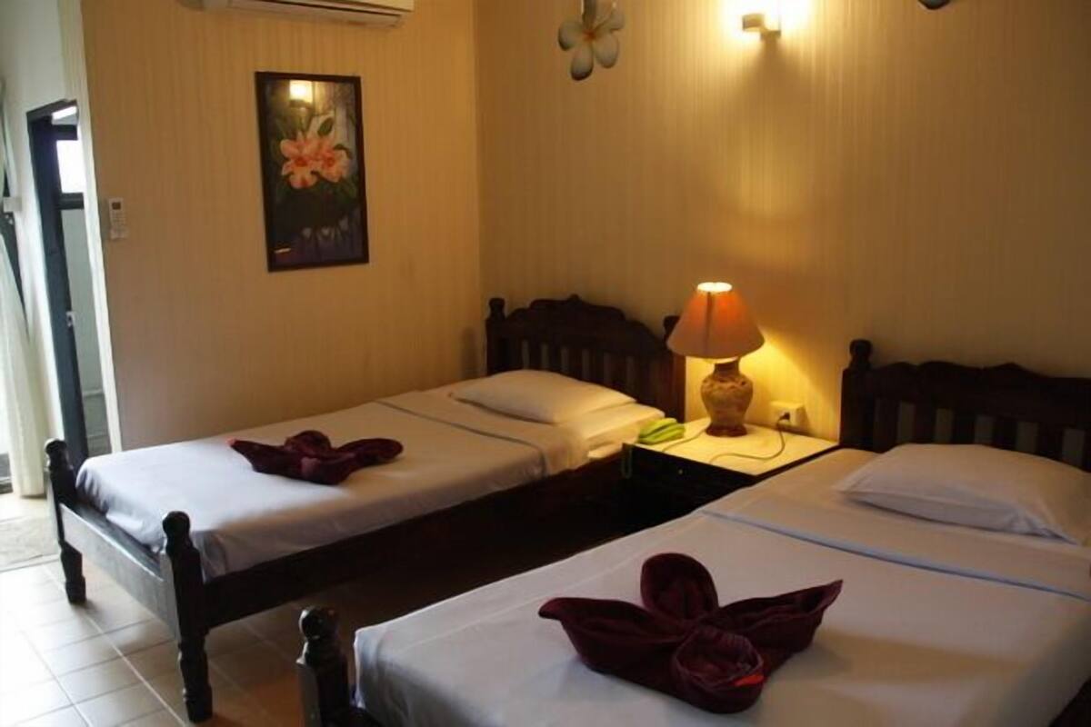 Quarto de hotel com duas camas de solteiro, uma mesa de cabeceira com abajur, um quadro de flores na parede e uma porta que dá para o exterior.