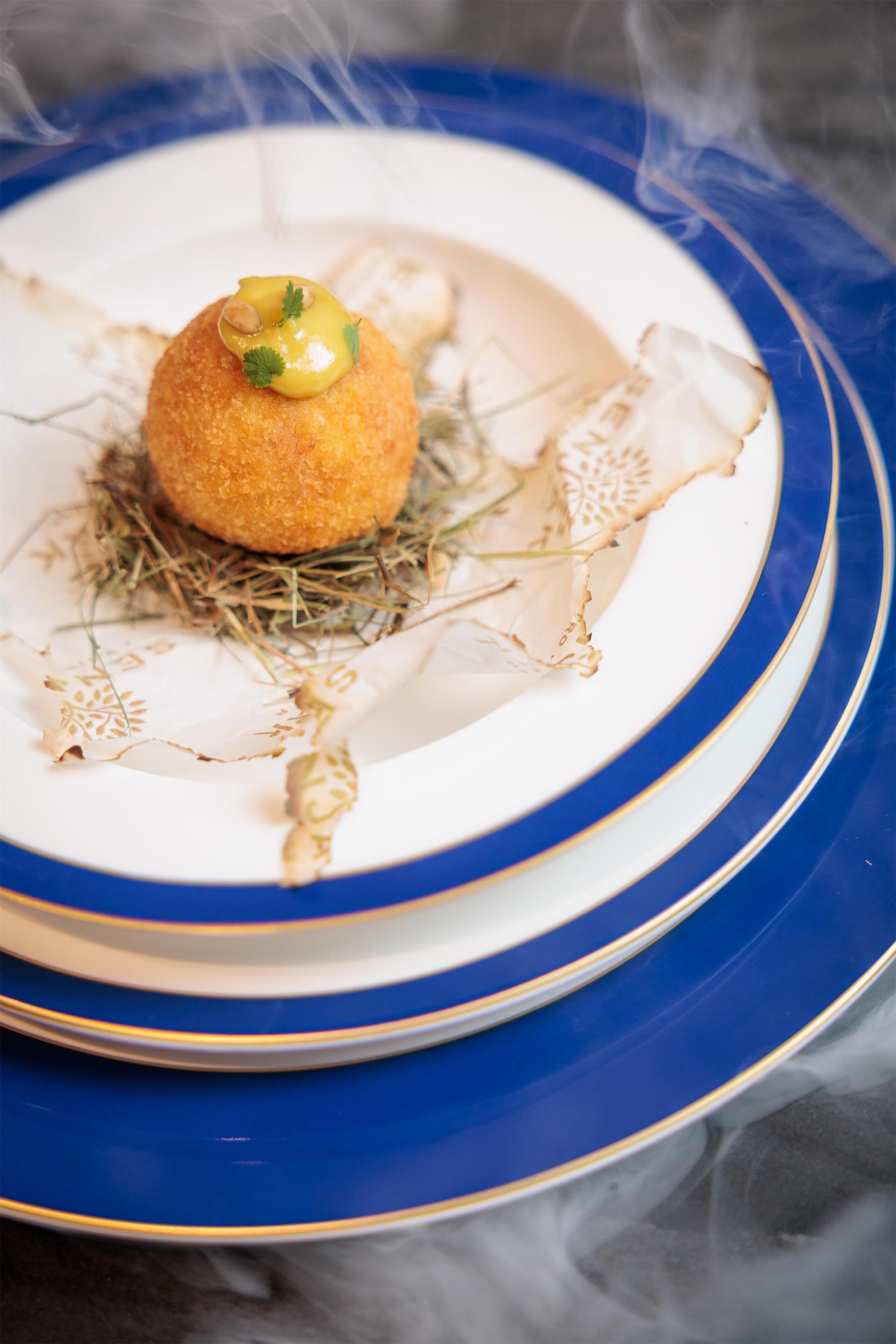 Prato com um alimento, possivelmente um croquete, decorado com ervas e molho, sobre um prato branco com borda azul.