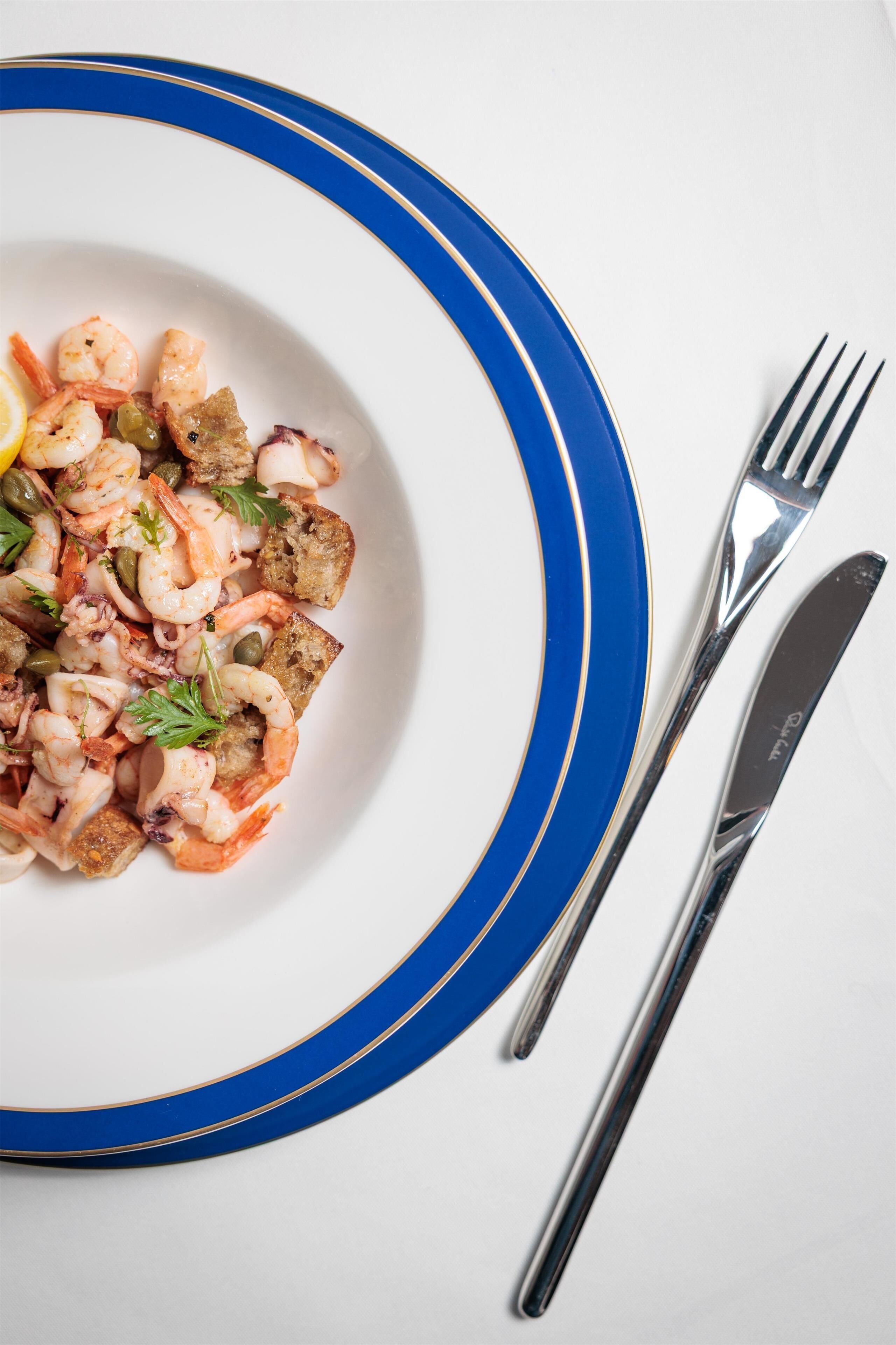 Uma salada de camarão e polvo com um limão, acompanhada de garfo e faca sobre uma mesa branca.