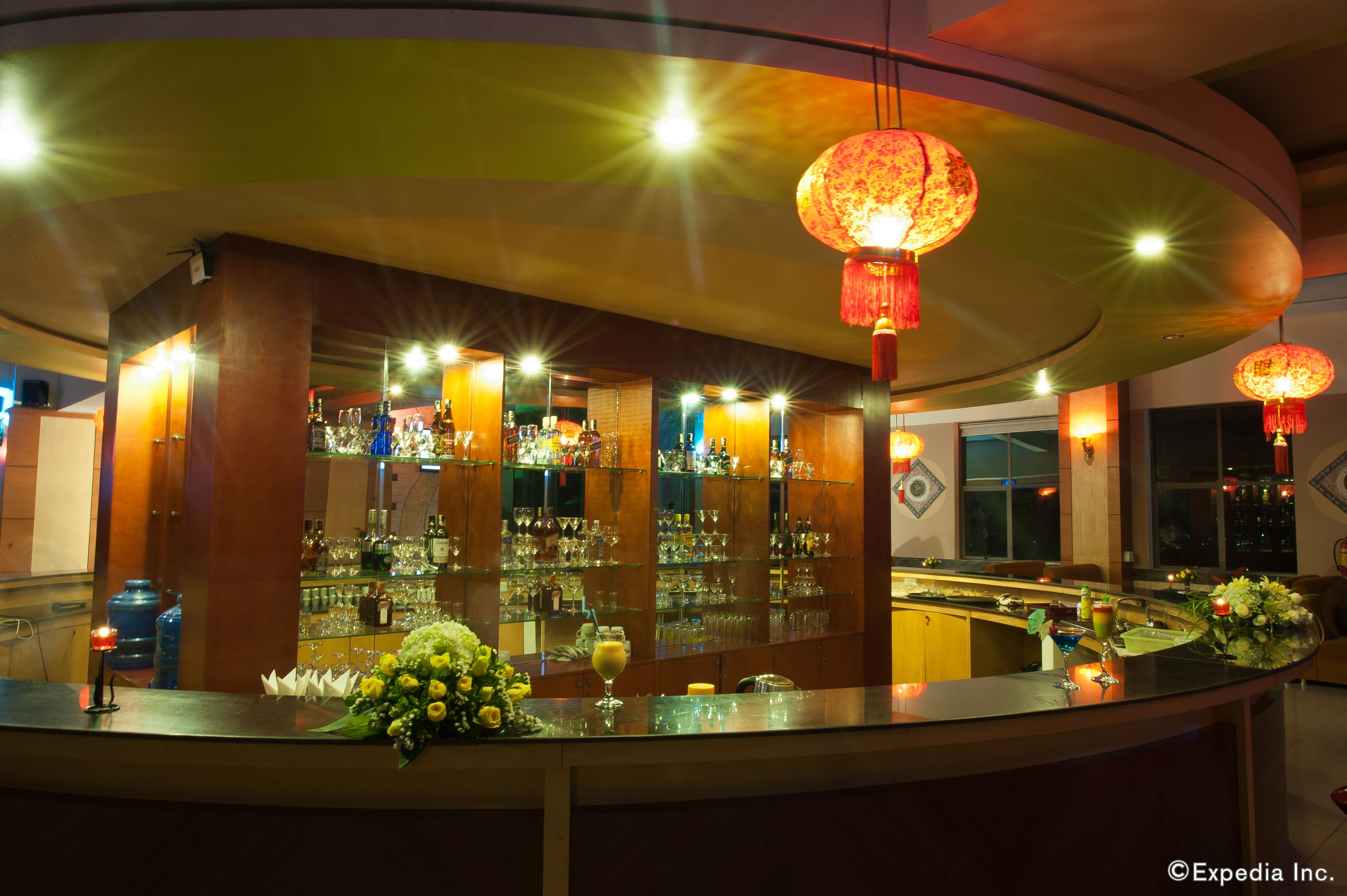 Uma área de bar com prateleiras de garrafas e copos, um balcão com arranjo de flores e lanternas penduradas.