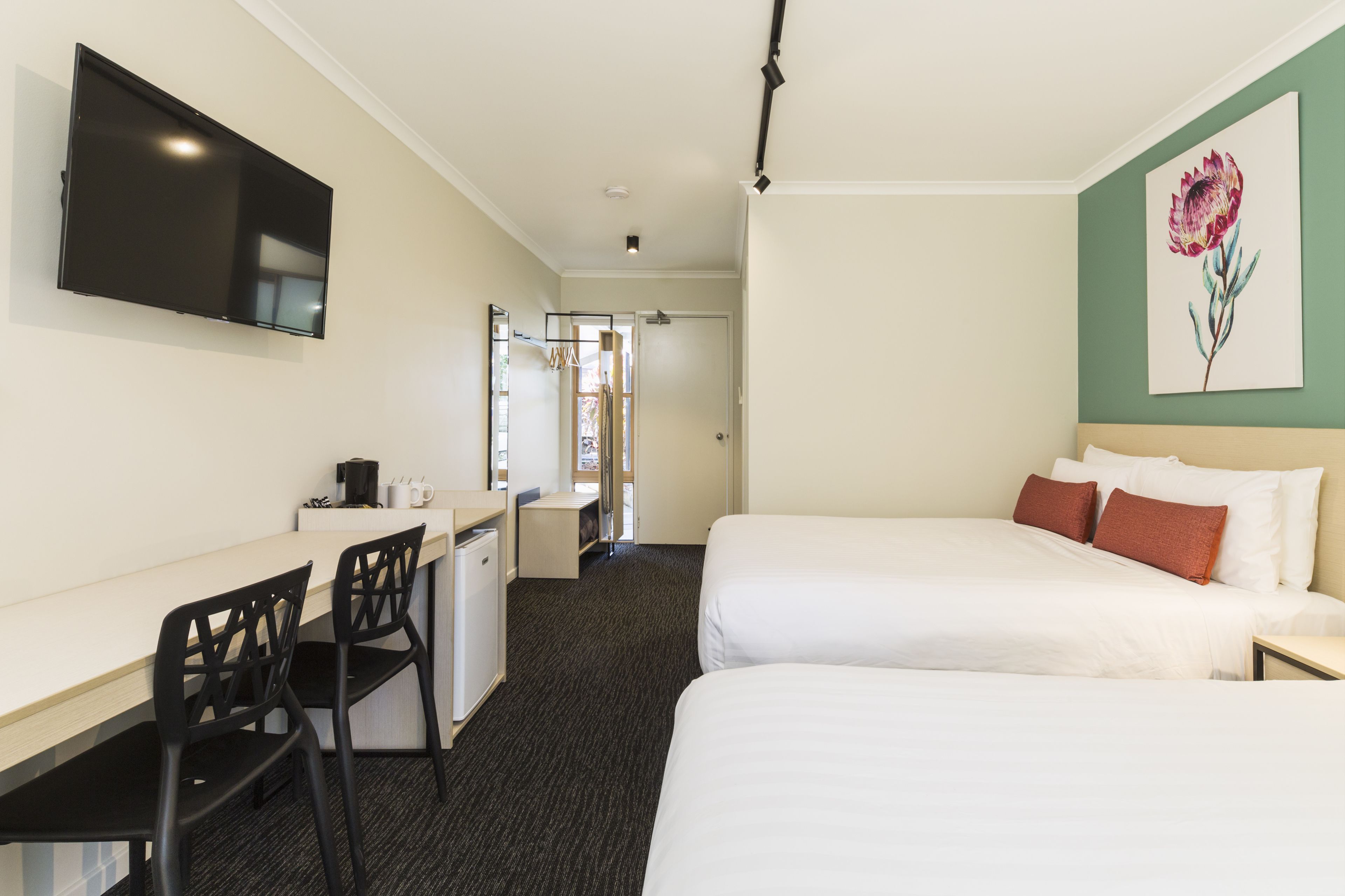 Quarto de hotel com duas camas, uma escrivaninha com cadeiras, uma televisão de tela plana e um quadro na parede.