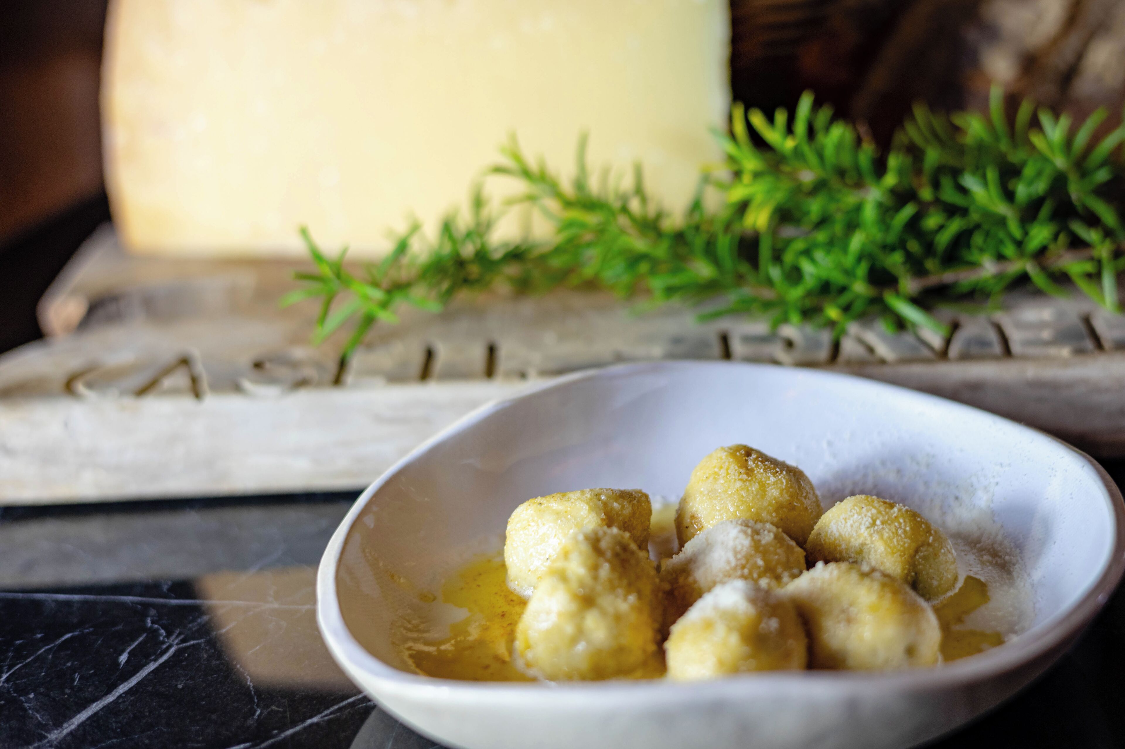 Uma tigela de comida com molho cremoso, um queijo inteiro e vegetação ao fundo.