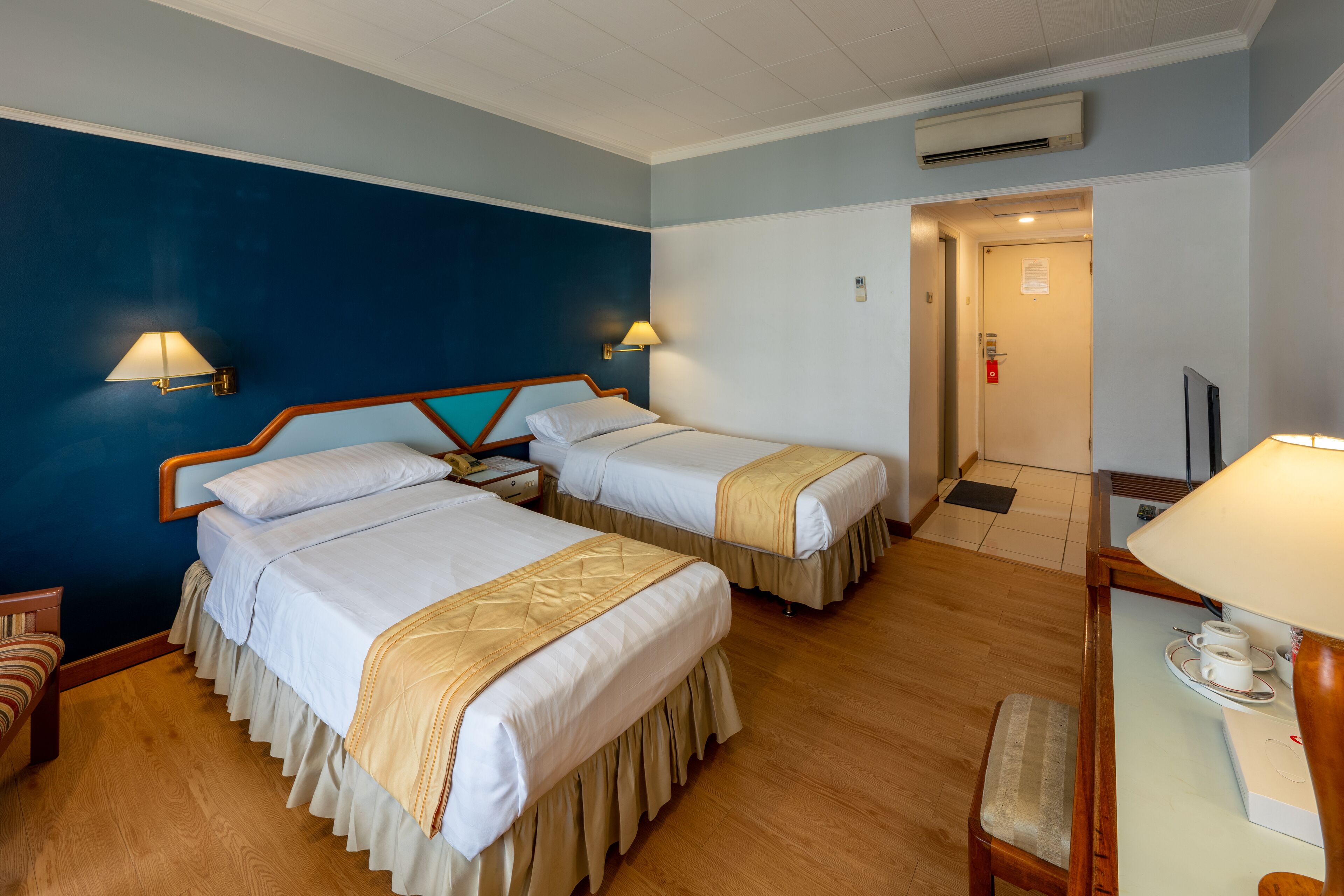 Quarto de hotel com duas camas, uma escrivaninha, uma cadeira, uma televisão e uma porta que dá para outro quarto.
