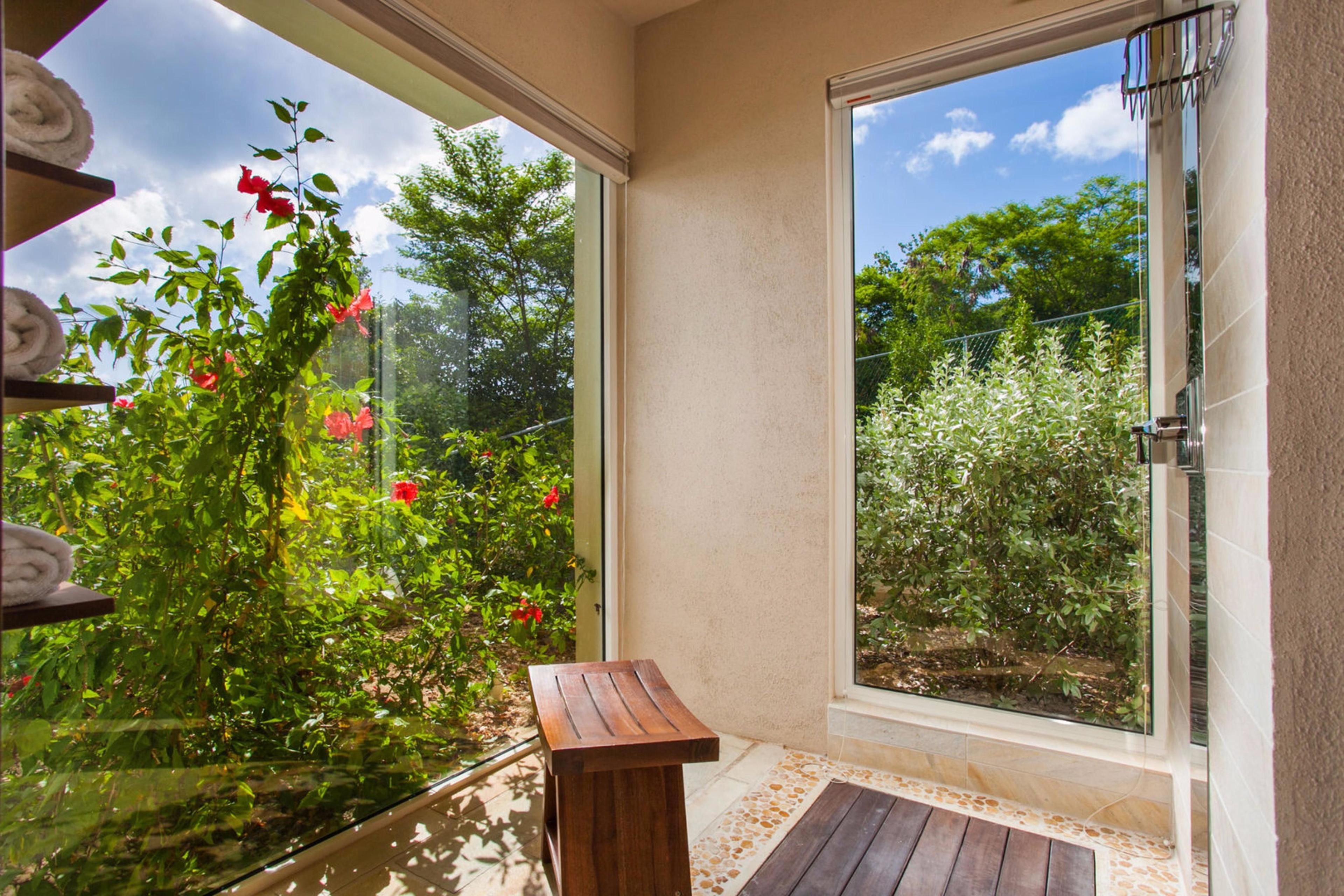 Varanda com mesa de madeira, porta de vidro e vista para um jardim com flores vermelhas.