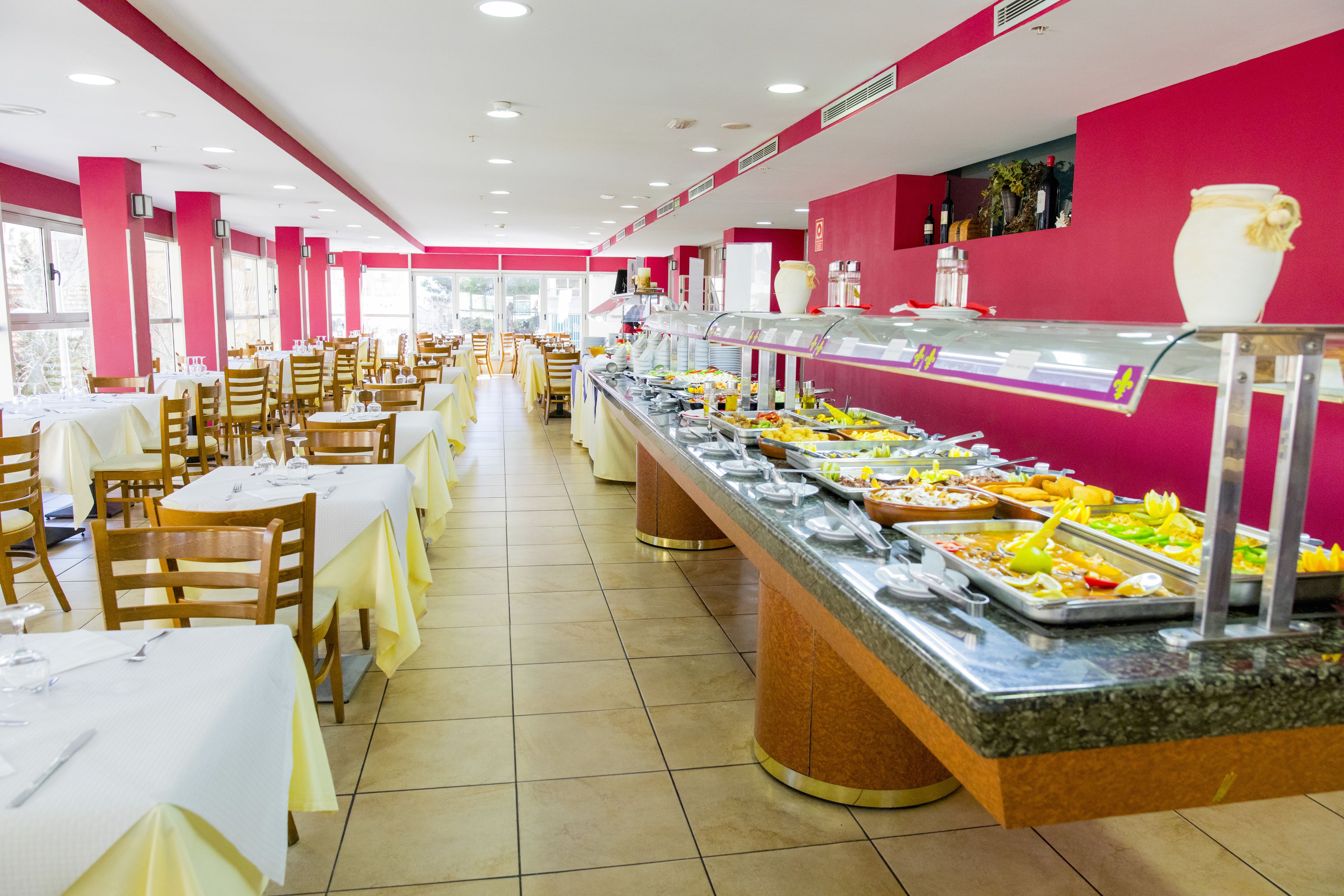 Uma área de buffet com diversas estações de comida, mesas com cadeiras e um interior claro, com paredes cor-de-rosa e grandes janelas.