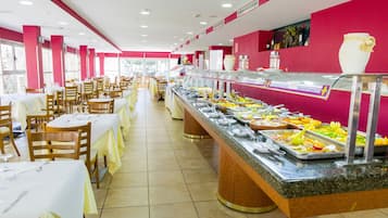 Uma área de buffet com diversas estações de comida, mesas com cadeiras e um interior claro, com paredes cor-de-rosa e grandes janelas.