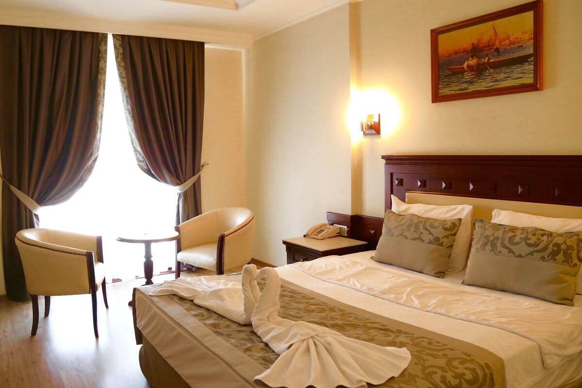 Quarto de hotel com cama, duas cadeiras, mesa de cabeceira com telefone e um quadro na parede.