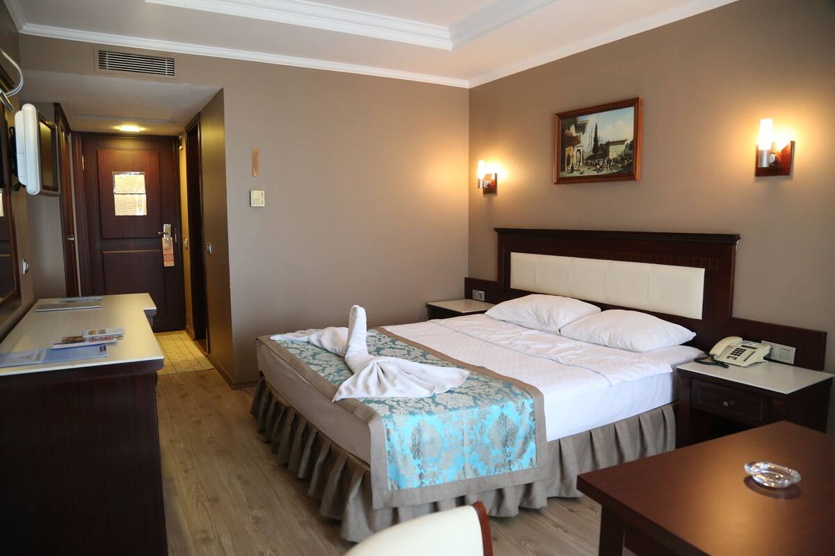 Quarto de hotel com cama, mesa de cabeceira e escrivaninha com telefone.
