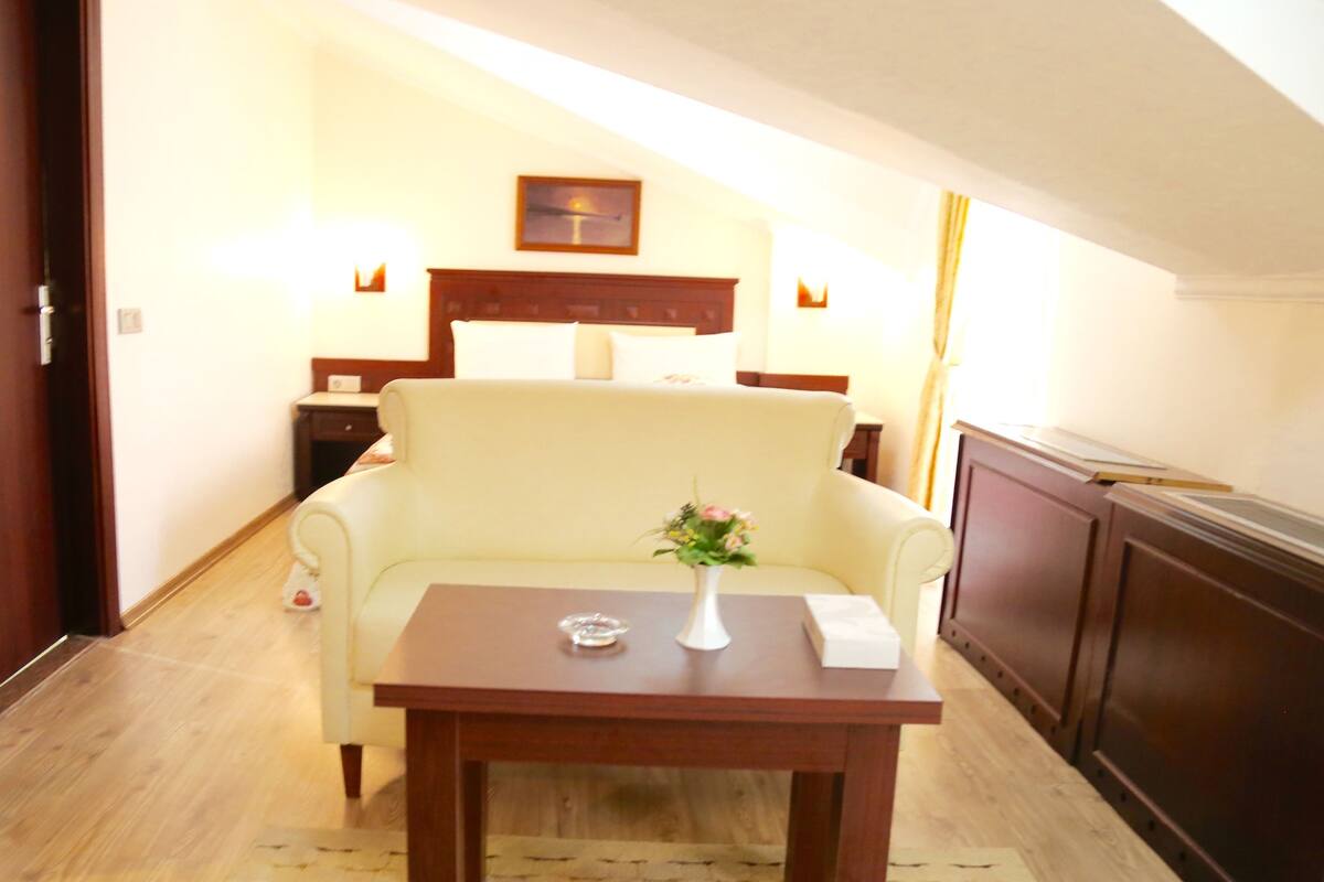 Quarto de hotel com sofá, uma mesinha com um vaso, mesa de cabeceira, abajur e um quadro na parede.