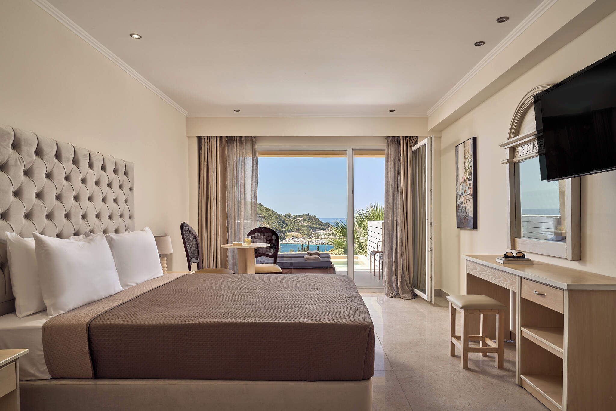 Quarto de hotel com uma cama grande, uma escrivaninha, uma cadeira, televisão e vista para o mar.