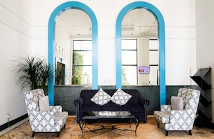 Um hall com um sofá azul escuro, poltronas estampadas, uma mesa de centro de vidro e um espelho grande com arcos azuis.