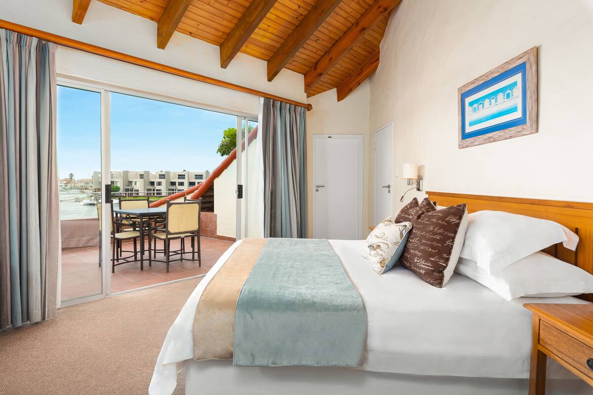 Quarto de hotel com cama, mesa de cabeceira, janela com cortinas, um quadro e vista para um edifício e uma árvore lá fora.