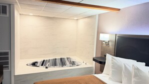 Quarto de hotel moderno com parede de mármore, uma banheira grande, cama com roupa branca e mesa de cabeceira com abajur.
