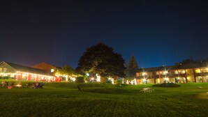 Pátio de hotel bem iluminado à noite, com gramado, uma grande árvore e vários edifícios com janelas iluminadas.