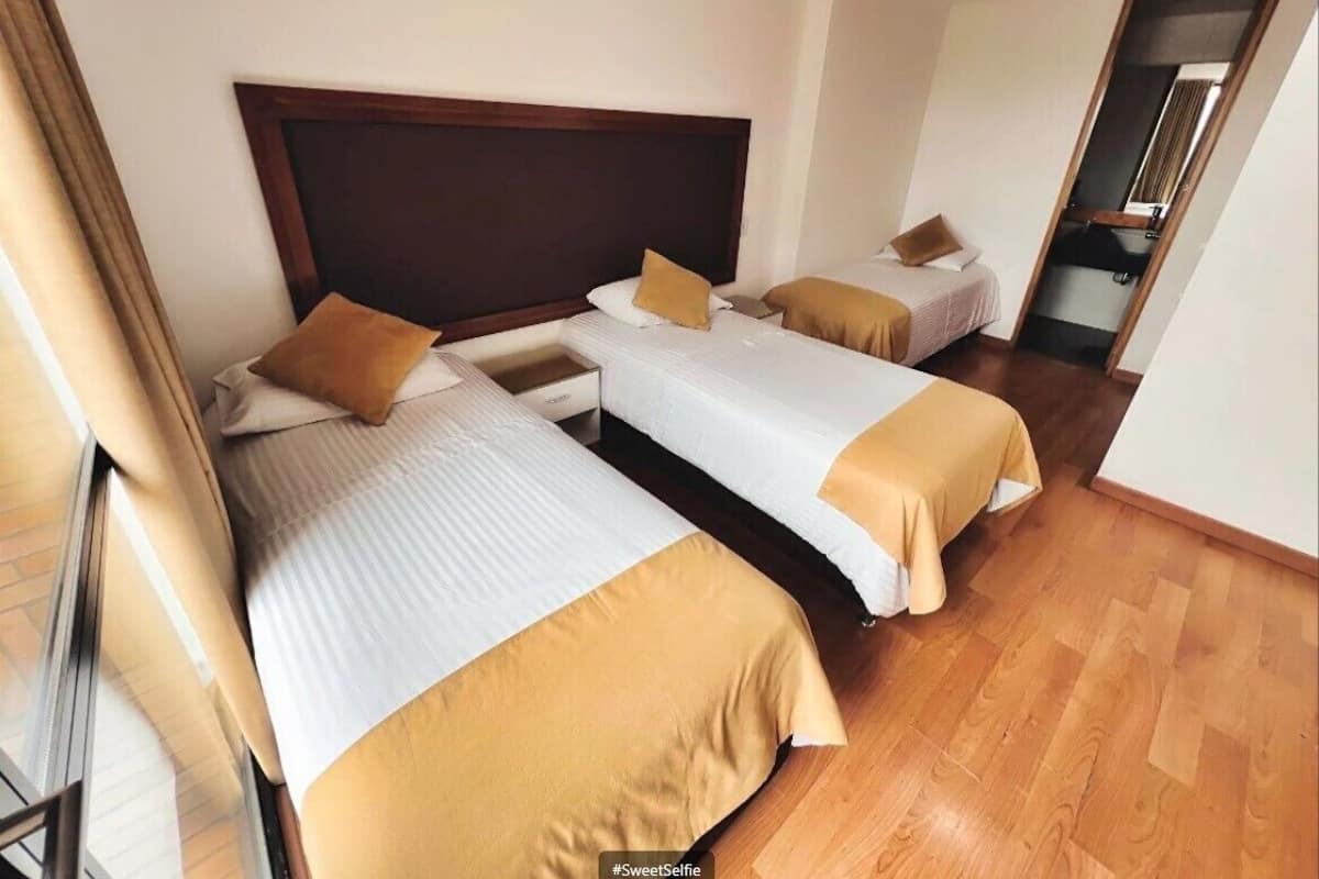 Quarto de hotel com três camas individuais, cada uma com cabeceira marrom e roupa de cama branca.