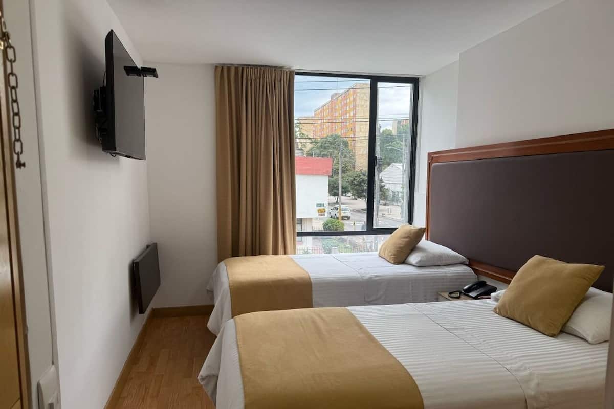 Quarto de hotel com duas camas, televisão presa à parede, janela com vista para edifícios e piso de madeira.
