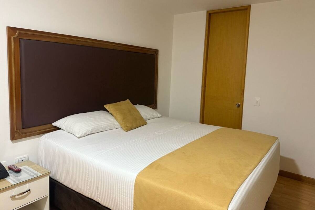Cama bem arrumada com edredom branco e manta marrom, cabeceira de madeira, mesa de cabeceira com abajur e porta de madeira.