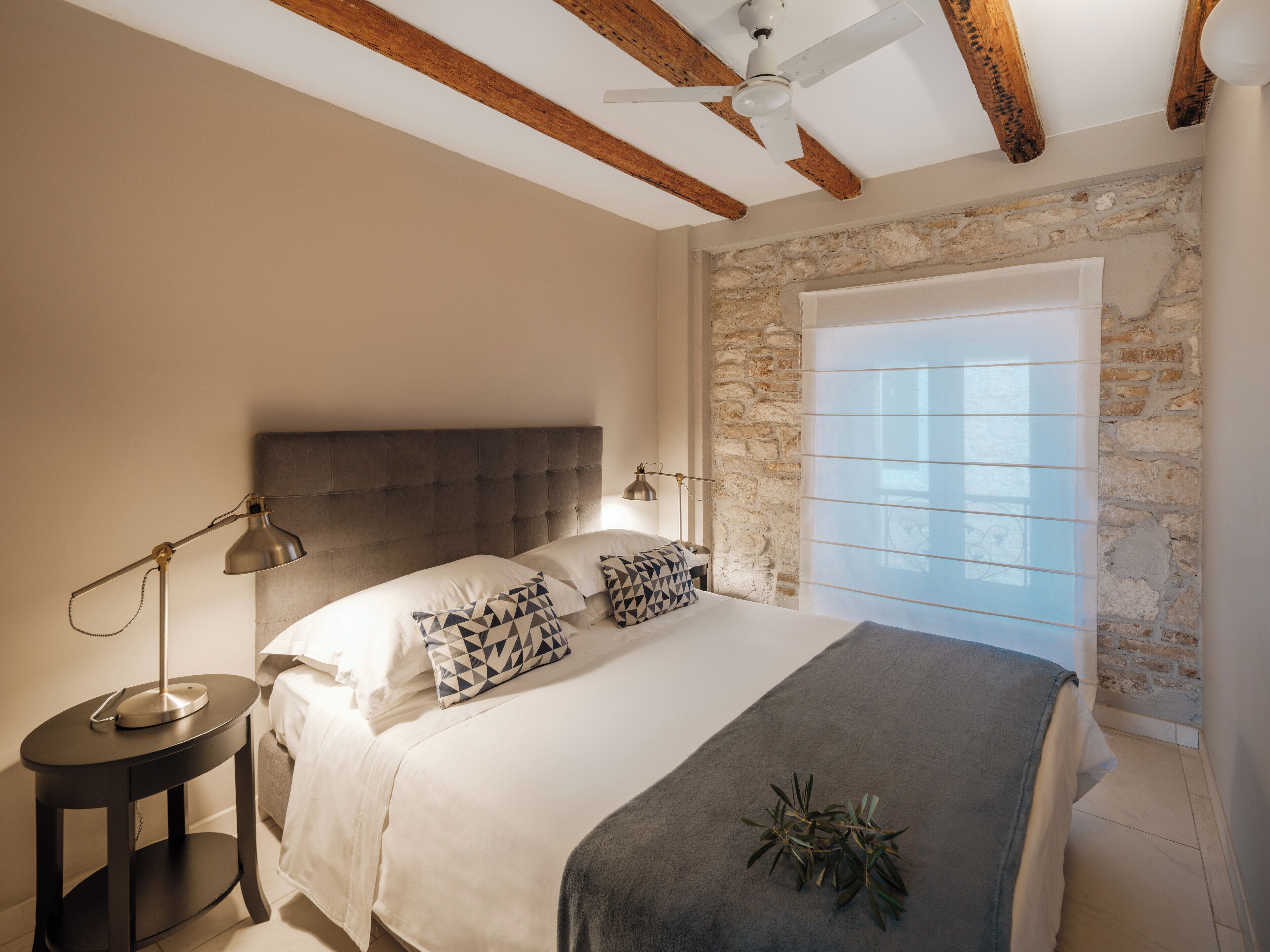 Um quarto com parede de pedra, uma cama com cobertor cinza e um ventilador de teto.