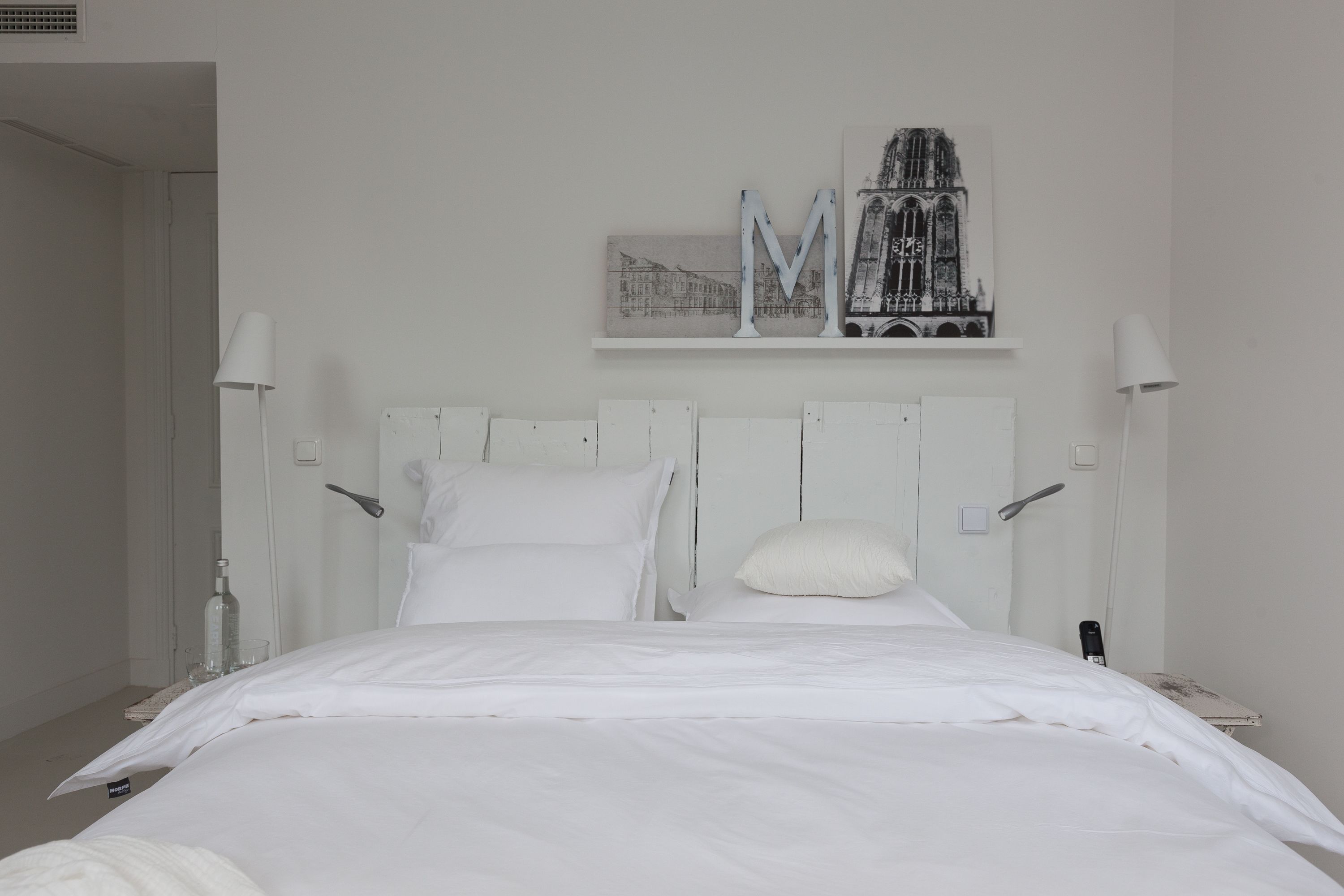 Quarto branco com cama, mesa de cabeceira, abajur e quadro de uma catedral na parede.