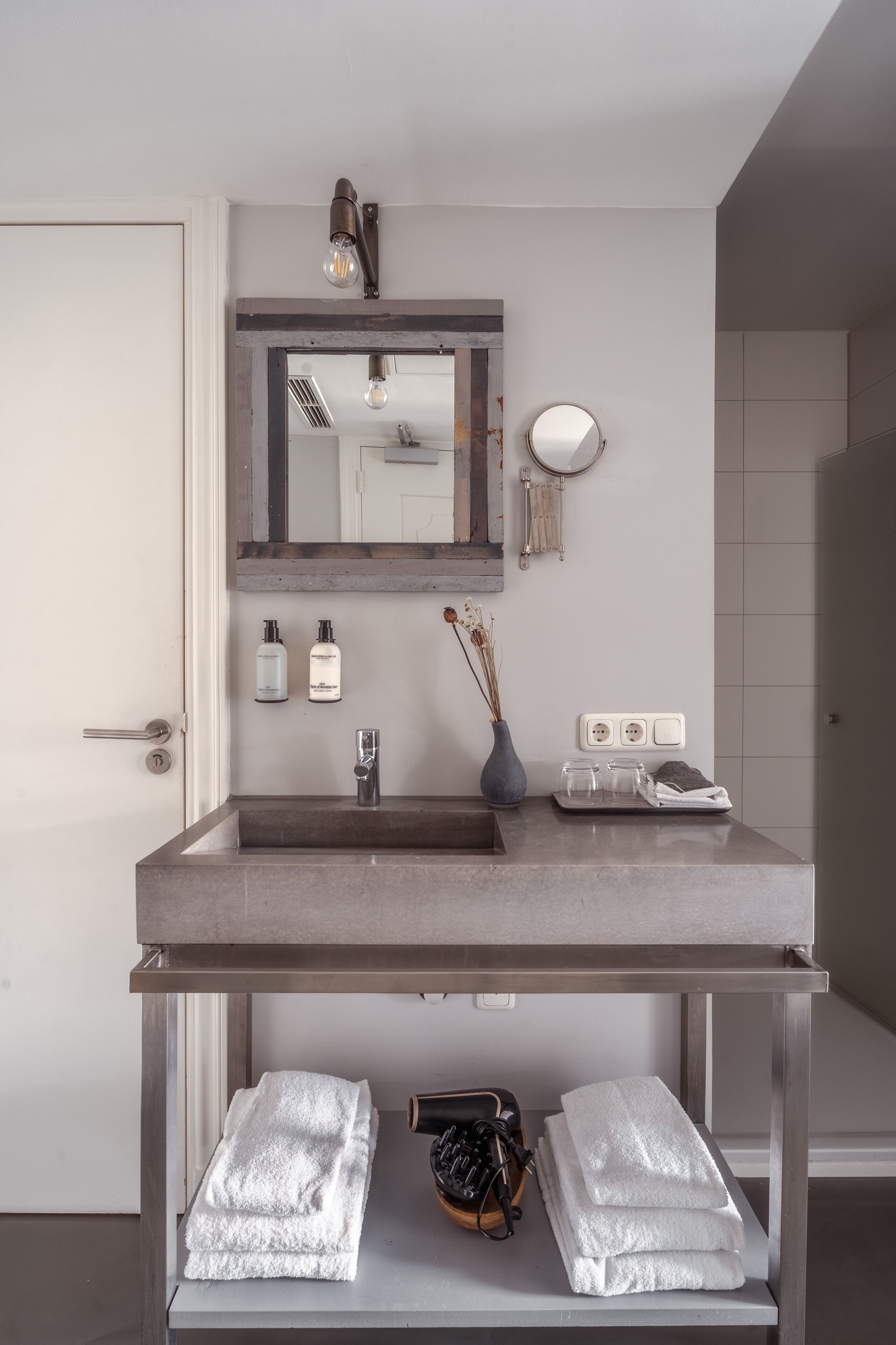 Casa de banho com lavatório cinzento, um espelho e um porta-toalhas. Existem duas toalhas no porta-toalhas, um vaso com flores secas e um frasco de champô.