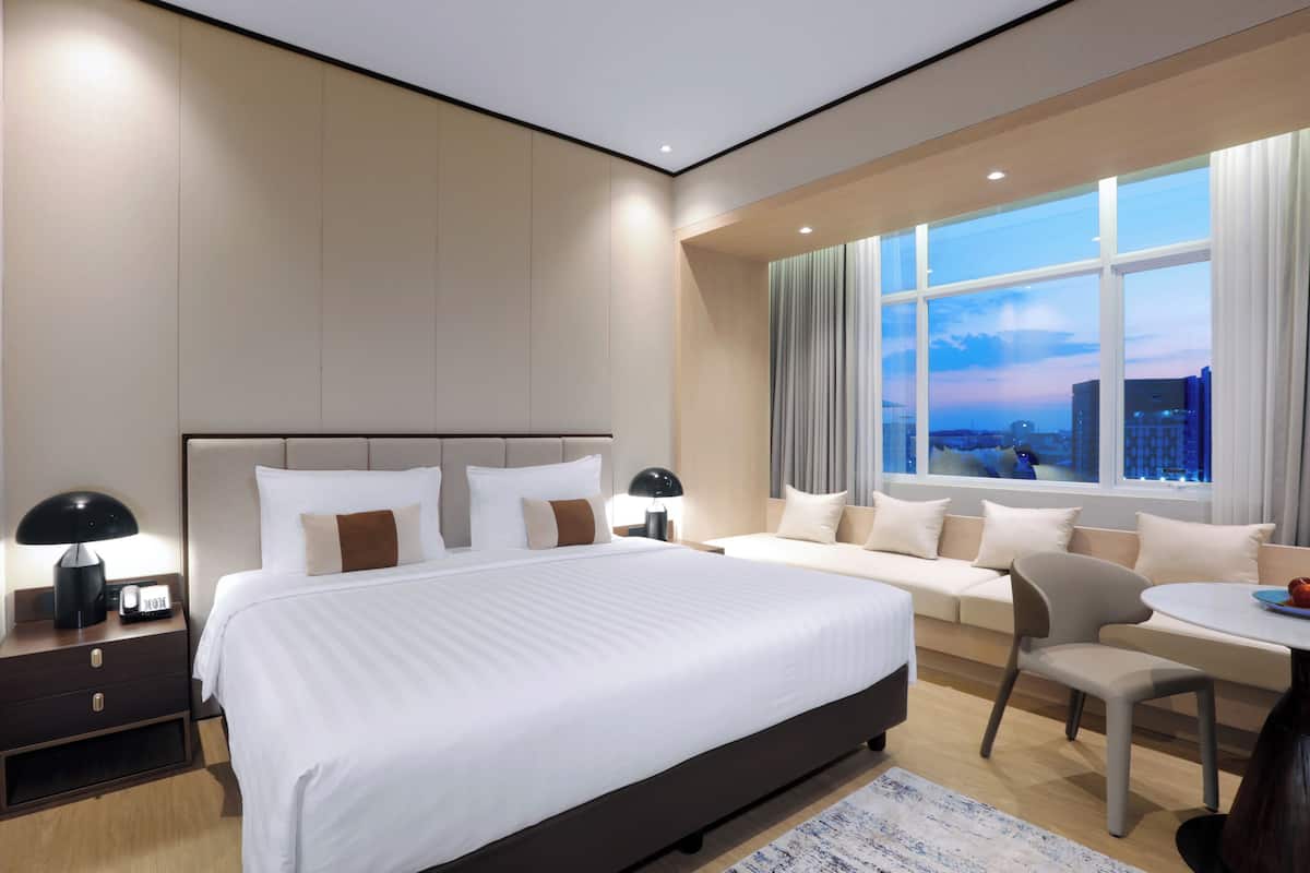Quarto de hotel moderno com uma cama grande, um sofá, uma mesa de jantar e vista para o horizonte urbano através de amplas janelas.