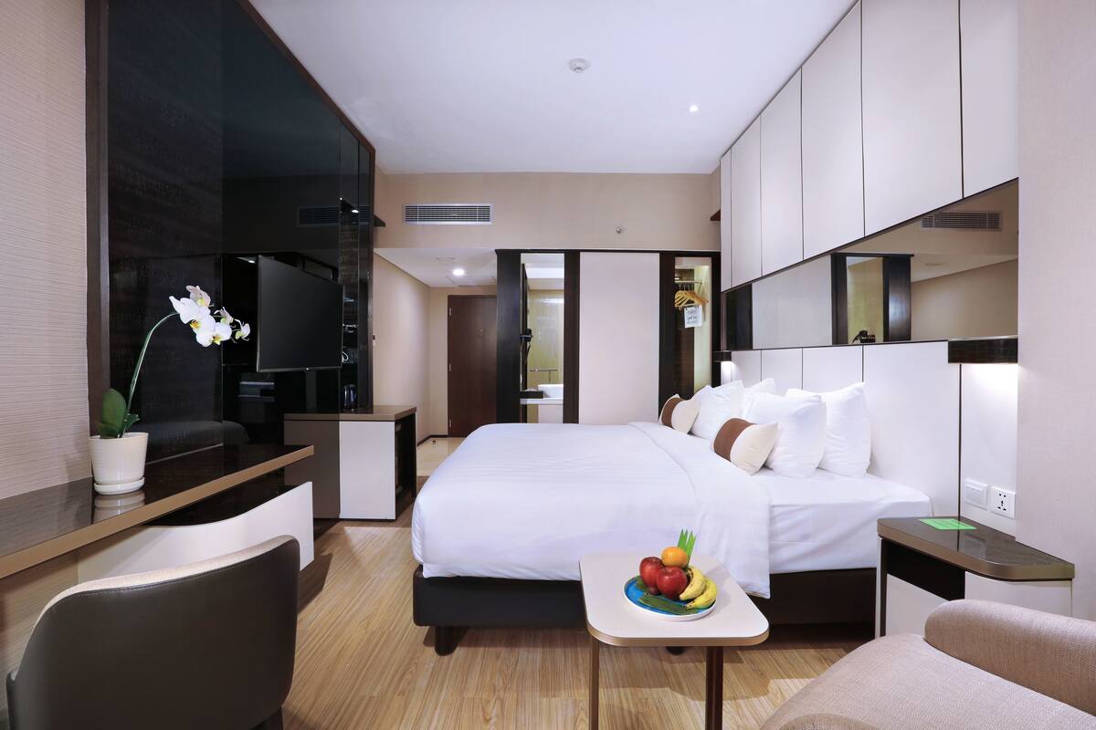 Quarto de hotel moderno com uma cama grande, uma escrivaninha com computador, uma mesinha com frutas e uma televisão sobre um suporte.