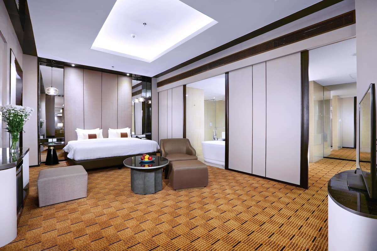 Quarto de hotel moderno com uma cama grande, área de estar com mesa de centro e banheiro separado.