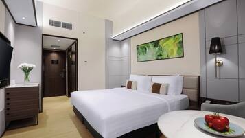 Quarto de hotel moderno com uma cama grande, uma mesa de cabeceira com um vaso, uma televisão de tela plana, uma escrivaninha com gavetas, uma cadeira, uma mesa redonda com uma travessa de fruta e um quadro na parede.