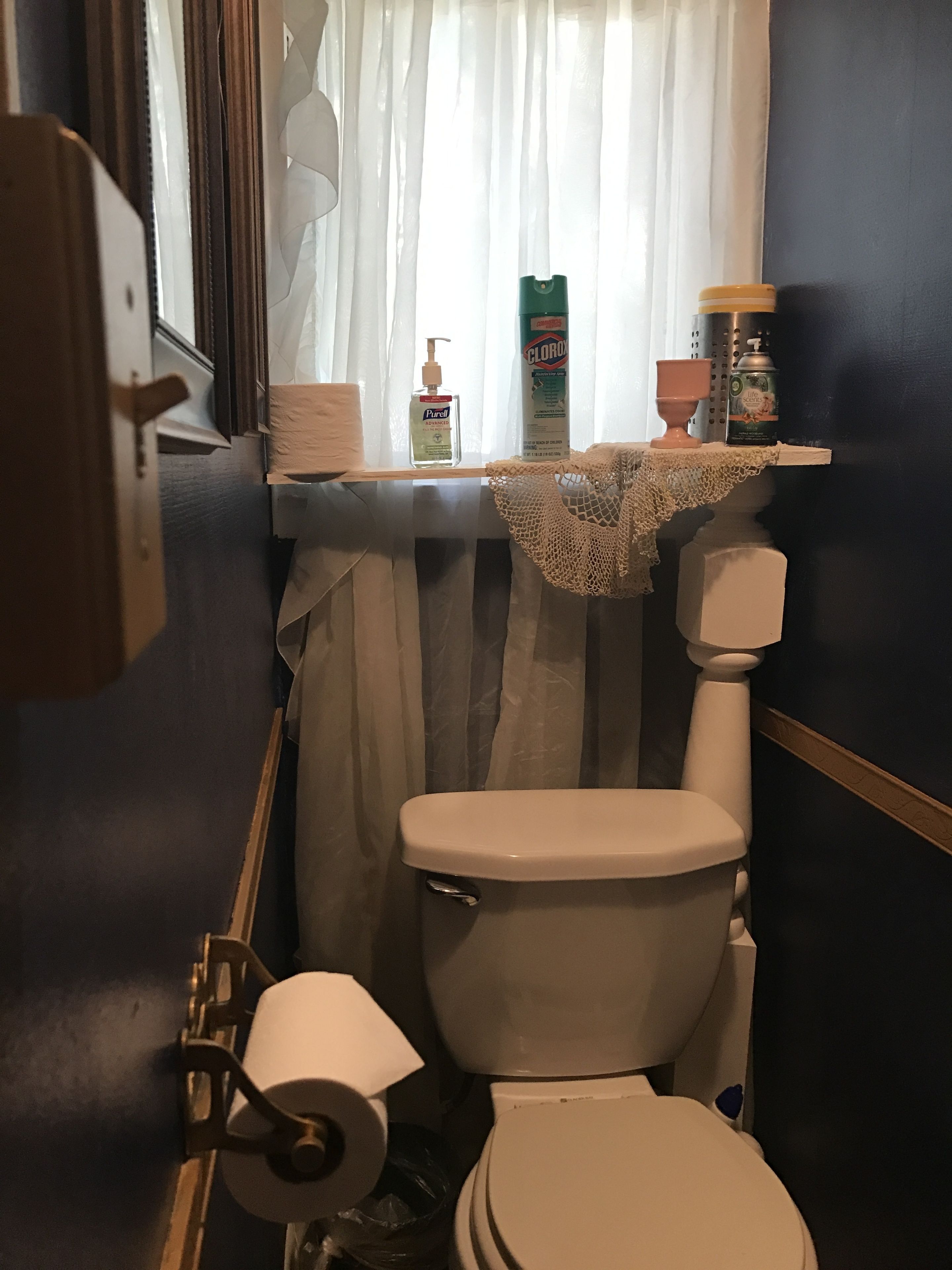 Um banheiro com vaso sanitário, uma prateleira com produtos de higiene, um rendado e uma janela com cortinas.