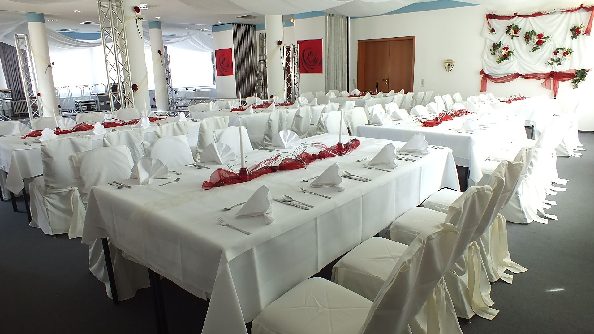 Uma sala de banquetes com fileiras de mesas preparadas para um evento formal, toalhas brancas e detalhes em vermelho.