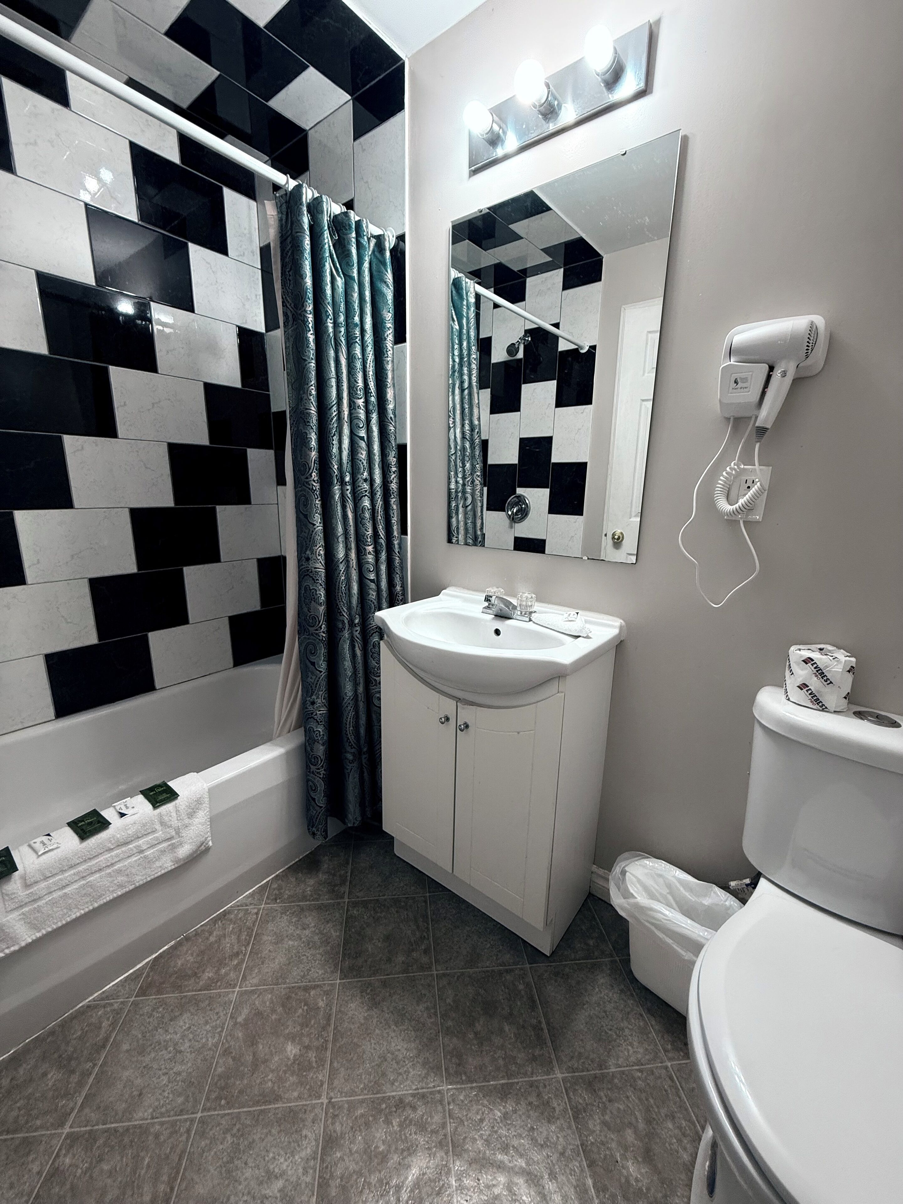 Um banheiro com parede de azulejos quadriculada em preto e branco, chuveiro com cortina, pia com espelho acima e vaso sanitário.