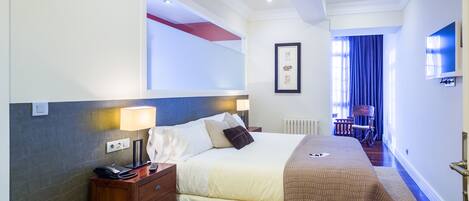 Quarto de hotel com cama, abajures, mesa de cabeceira de madeira, quadro na parede e vista para o corredor.