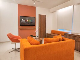 Uma área de recepção moderna com mobiliário laranja, uma televisão exibindo 'Zuri Express Lippo Cikarang' e uma porta de madeira.