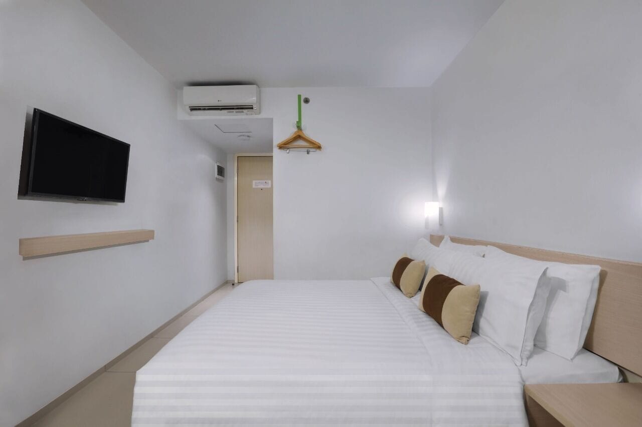 Quarto de hotel com uma cama, televisão fixada na parede, aparelho de ar condicionado e mesa de cabeceira de madeira.