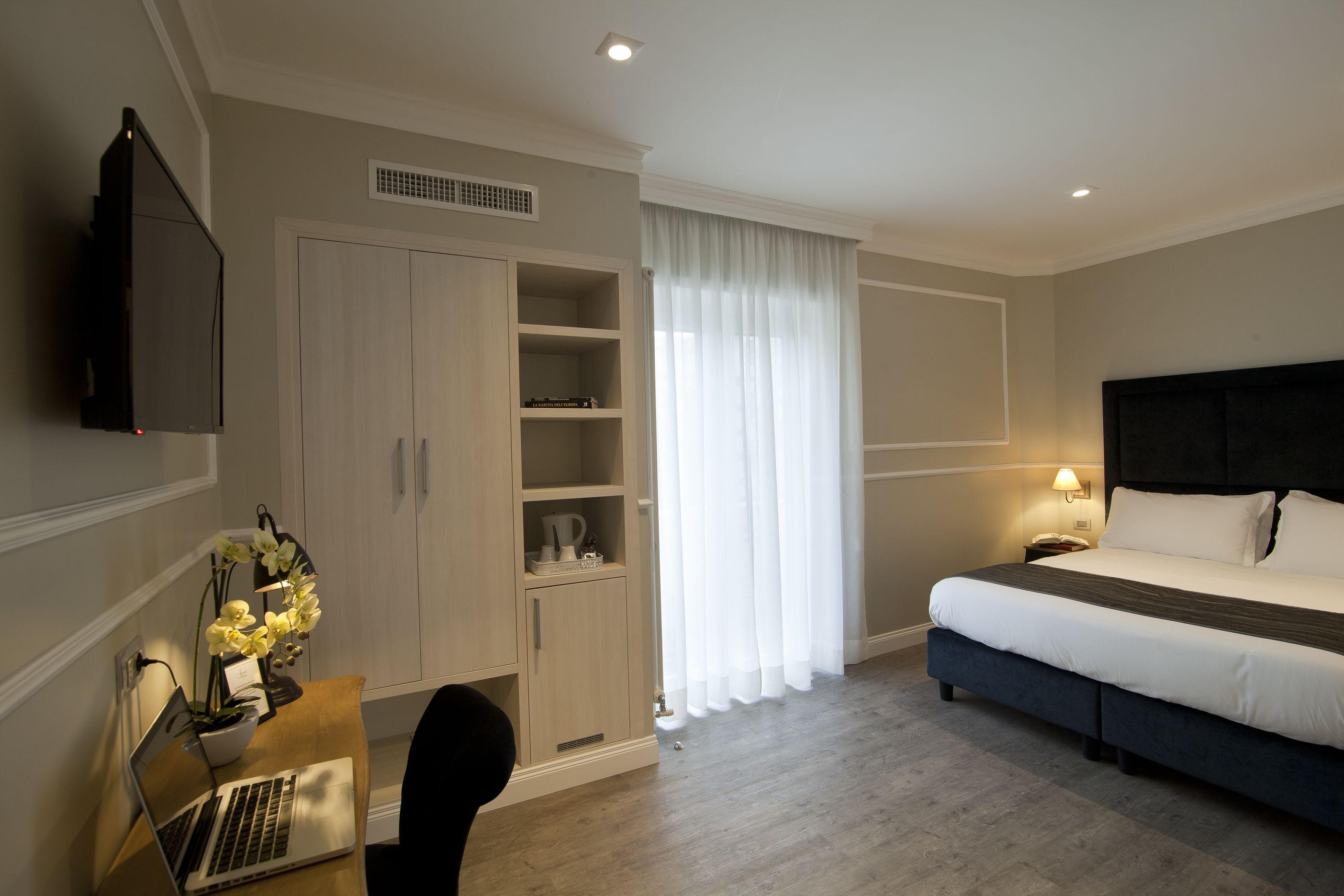 Quarto de hotel com cama, escrivaninha com laptop, guarda-roupa e televisão.