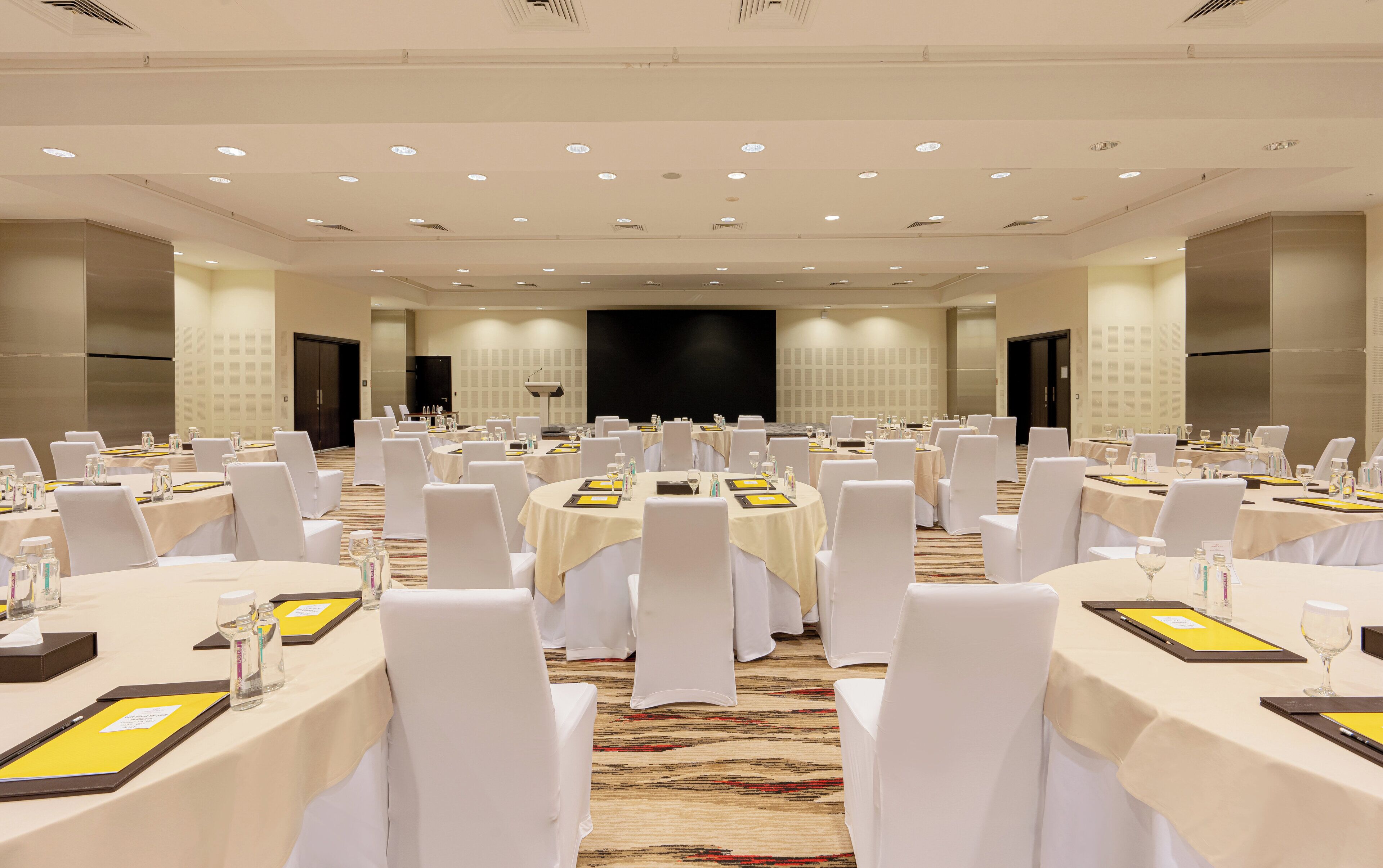 Sala de conferência com várias mesas redondas, cadeiras brancas e palco com fundo preto.