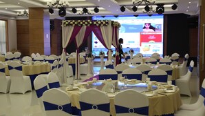Uma sala de banquetes com mesas redondas dispostas para um evento formal, com cadeiras brancas e fitas azuis, e um palco com um painel de flores.