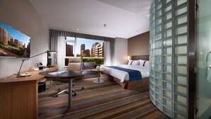 Quarto de hotel com cama, escrivaninha, cadeira, mesinha e vista para a paisagem urbana.