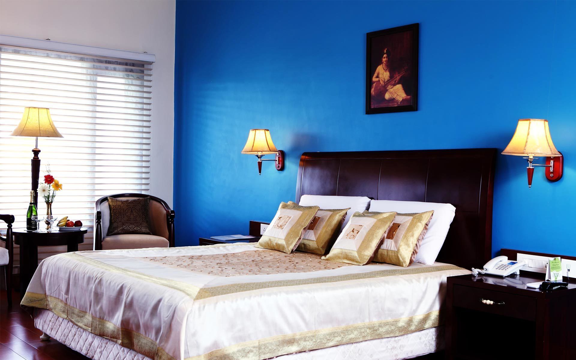 Quarto de hotel com uma cama grande, duas luminárias de cabeceira, uma cadeira, uma mesa com abajur e um quadro na parede.