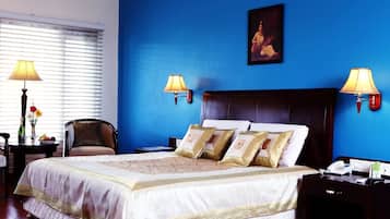 Quarto de hotel com uma cama grande, duas luminárias de cabeceira, uma cadeira, uma mesa com abajur e um quadro na parede.