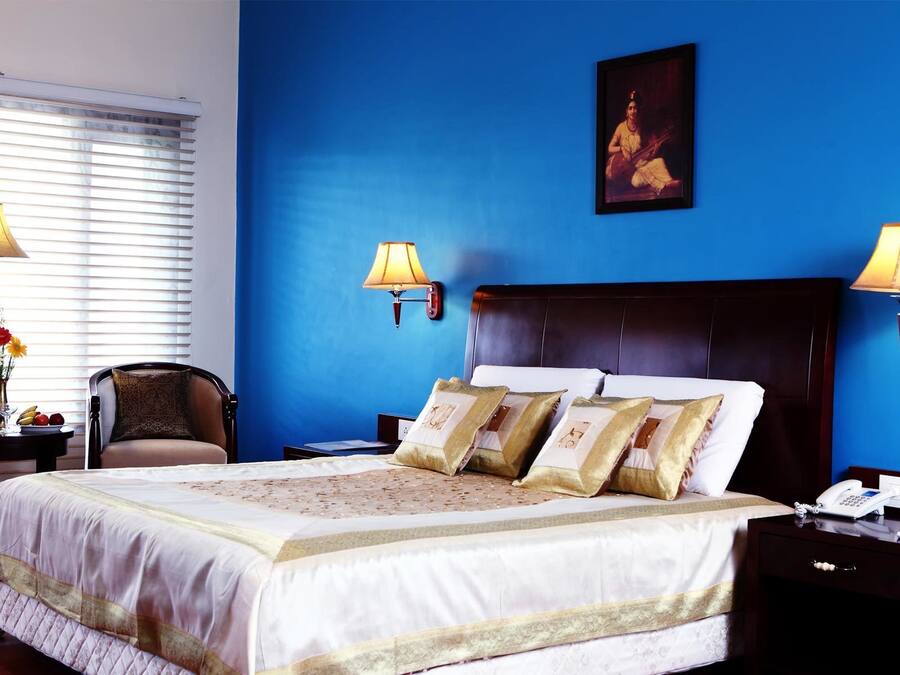 Quarto de hotel com uma cama grande, duas luminárias de cabeceira, uma cadeira, uma mesa com abajur e um quadro na parede.