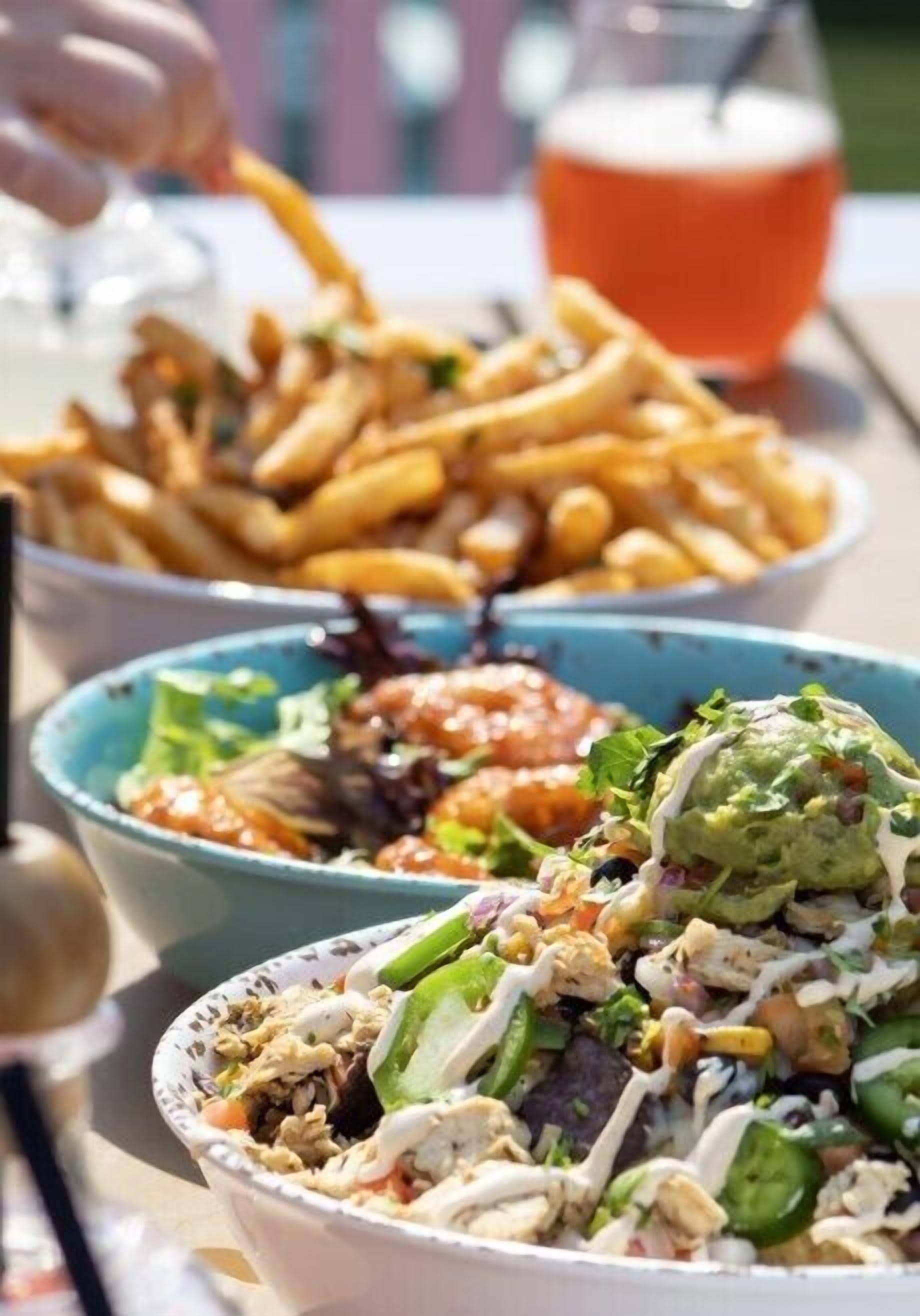 Uma mesa com três tigelas de comida: uma com salada, abacate e molho cremoso, outra com um prato misto, e uma terceira com batatas fritas e uma bebida ao fundo.